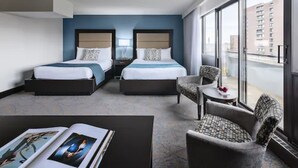 Quarto de hotel com duas camas, uma mesinha, uma cadeira e uma janela grande com vista para a cidade.