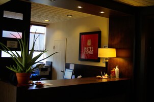 Uma receção de hotel com uma planta em vaso, um letreiro emoldurado com a inscrição "HOTEL L'EXPRESS", uma luminária de mesa e um frasco de álcool em gel.