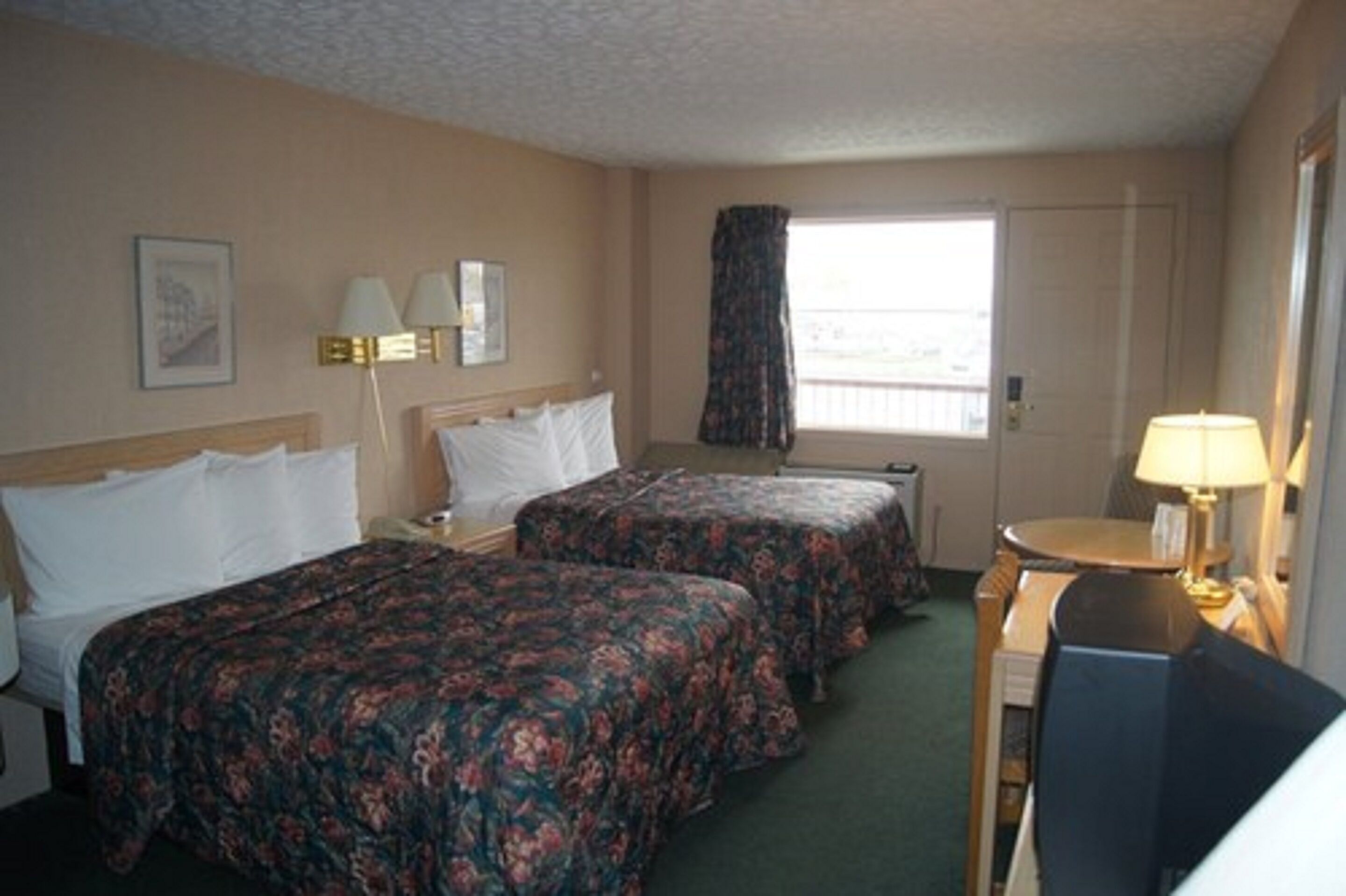 Quarto de hotel com duas camas, uma escrivaninha, uma cadeira, um abajur, uma janela com cortina e um quadro na parede.
