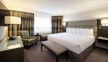 Quarto de hotel com uma cama grande, uma escrivaninha com luminária, uma cadeira, uma pequena mesa com um vaso e uma mesinha de cabeceira com relógio.
