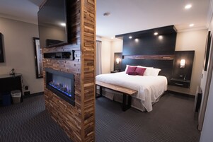 Quarto de hotel moderno com lareira, cama com roupa de cama branca, um banco e uma televisão de tela plana fixada em uma parede de madeira.