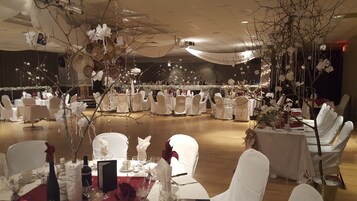Salão de banquetes com mesas redondas e cadeiras brancas, decorado com arranjos florais e luzes de fada.