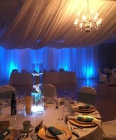 Salão de festas com mesas montadas para um evento formal, com um lustre e cortinas drapeadas.