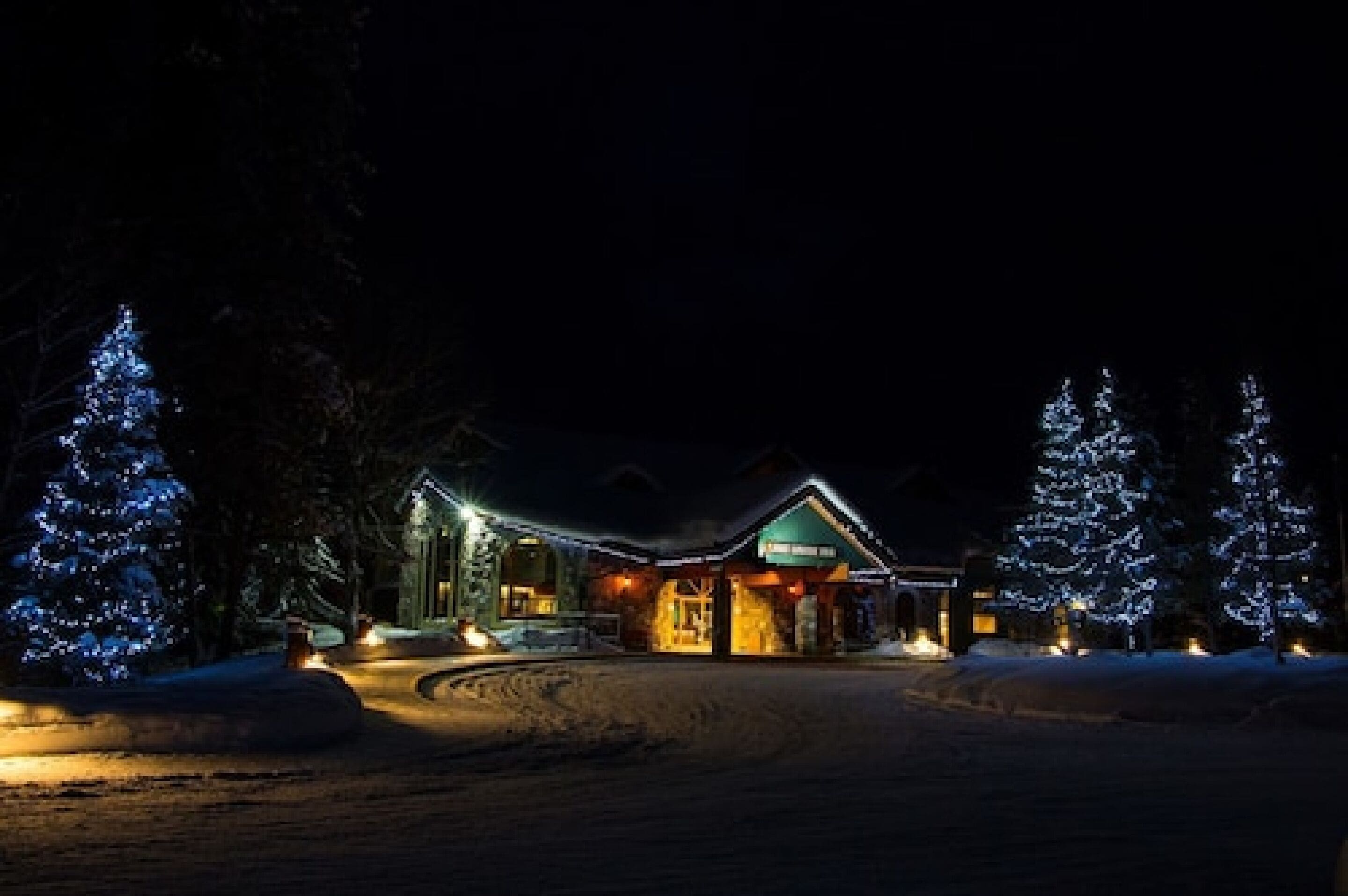 Paisagem coberta de neve com um edifício com telhado verde e janelas iluminadas, cercado por árvores carregadas de neve.