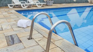 Piscina com deck revestido, escada de metal e cadeiras de descanso brancas ao fundo.