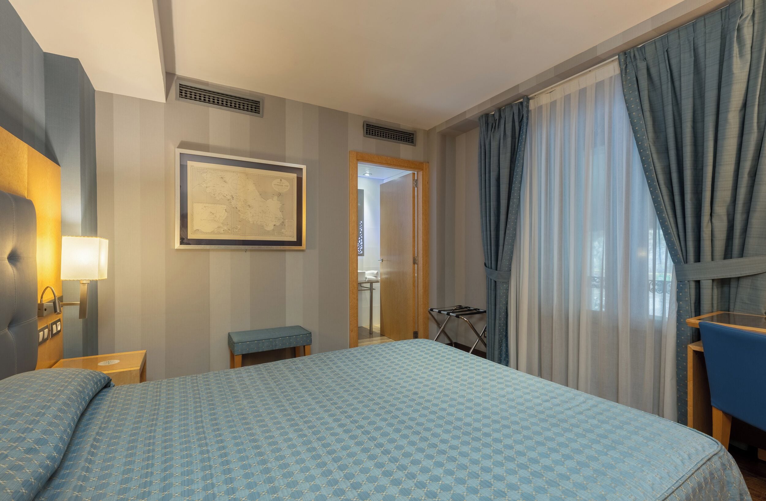 Quarto de hotel com uma cama, escrivaninha, cadeira, mesa pequena, abajur, espelho e janela com cortinas.