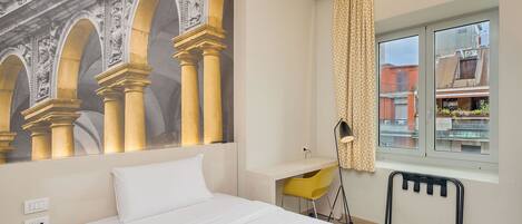 Quarto de hotel moderno com cama, escrivaninha, cadeira, luminária e uma grande janela com vista para prédios.