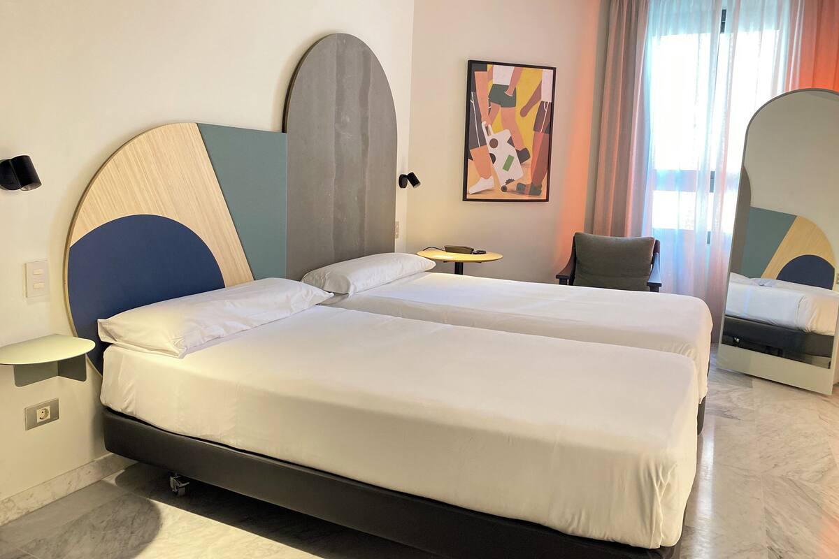 Quarto de hotel com duas camas, uma mesa redonda pequena, uma cadeira e um quadro na parede.
