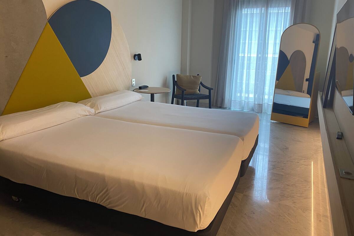 Quarto de hotel com uma cama grande, uma mesinha, uma cadeira e um espelho.