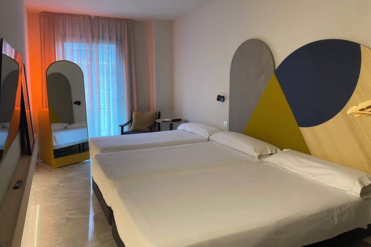 Quarto de hotel com duas camas, uma cadeira, uma mesinha e um espelho grande.