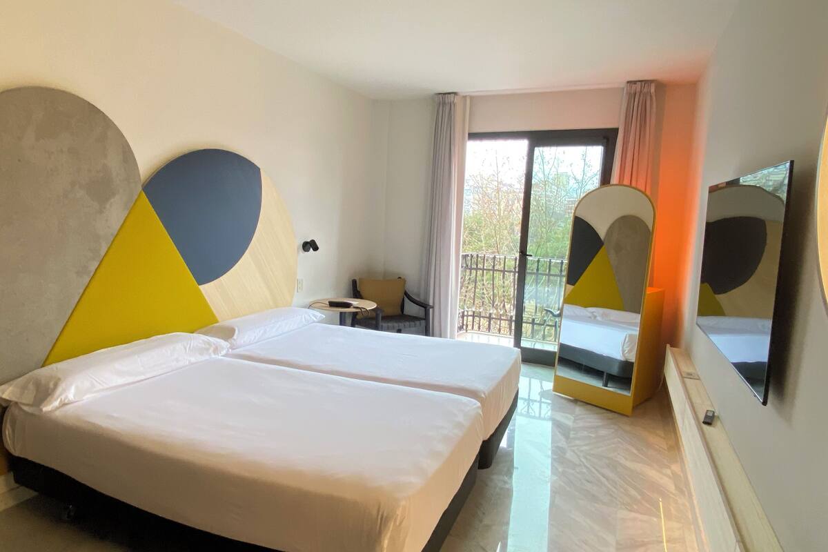 Quarto de hotel com duas camas, uma mesinha, uma cadeira e varanda com vista.