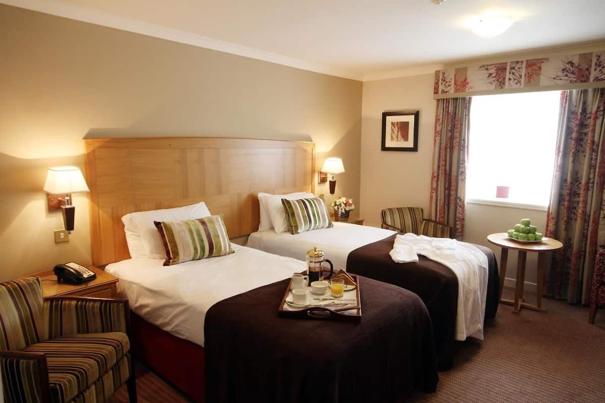 Quarto de hotel com duas camas, uma mesa pequena com itens de café da manhã, uma cadeira e uma janela com cortinas.