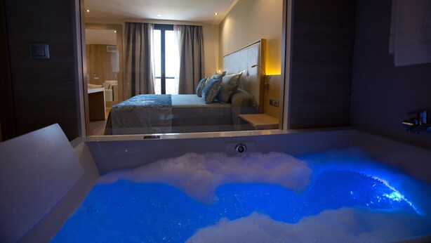 Quarto de hotel moderno com uma cama grande, banheiro separado e hidromassagem com iluminação azul.