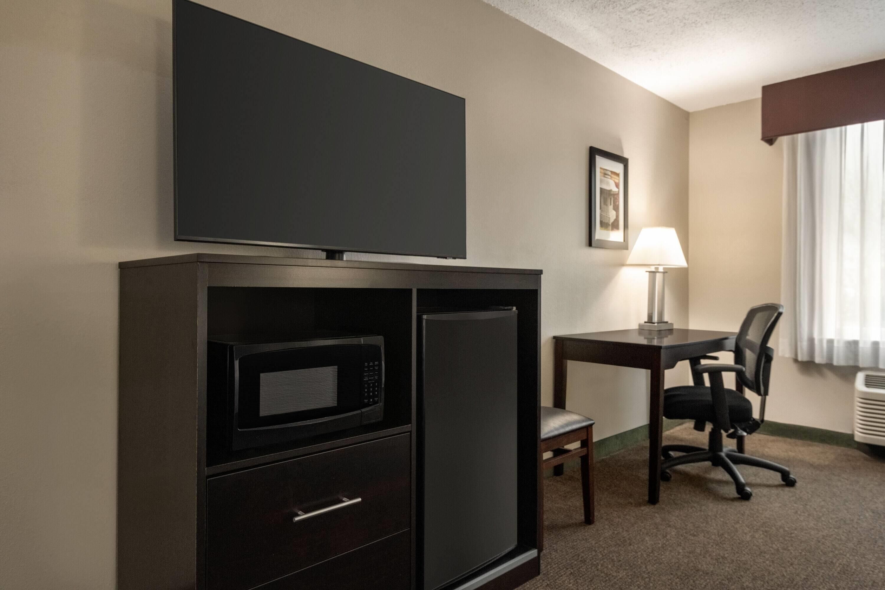 Quarto de hotel com televisão de tela plana, micro-ondas, geladeira, escrivaninha com cadeira, abajur e janela com cortinas.