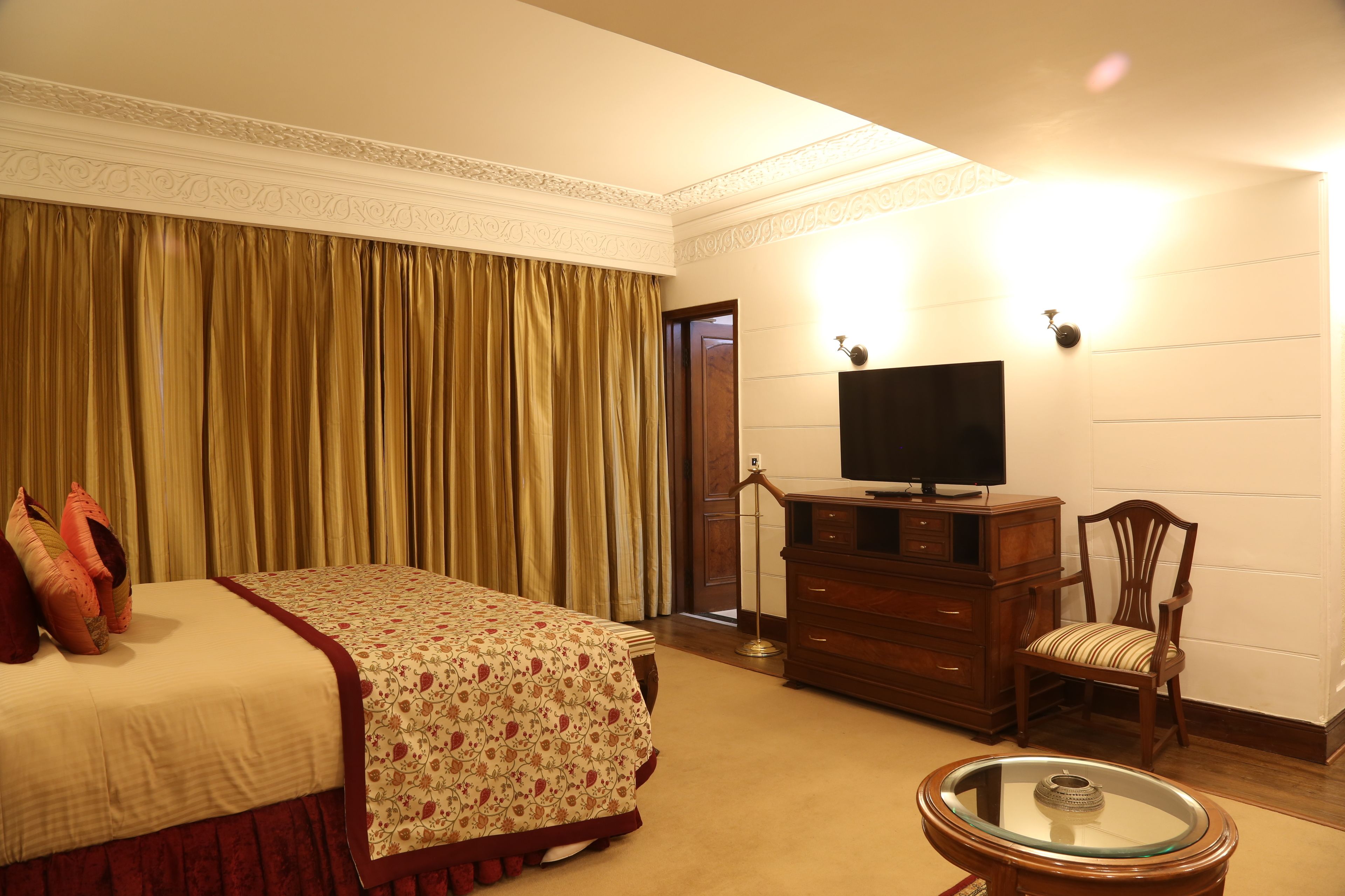 Quarto de hotel com uma cama, televisão, cómoda de madeira, cadeira e uma mesa pequena com tampo de vidro.