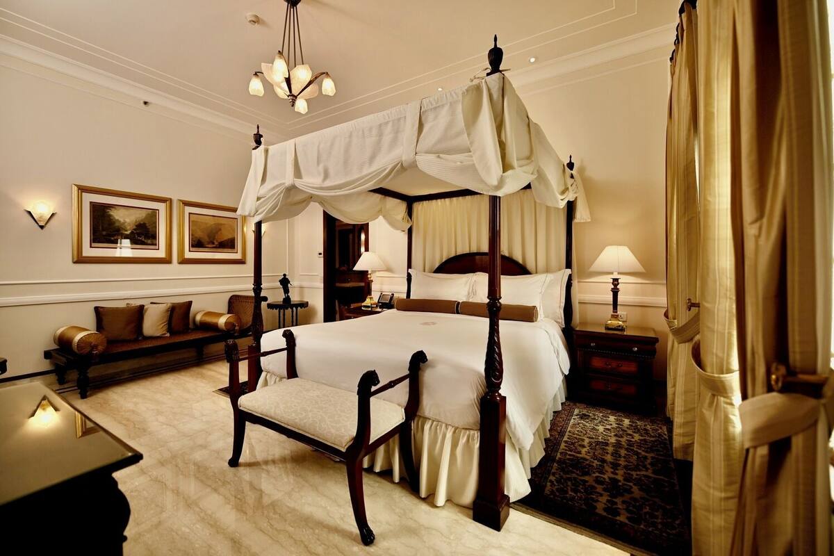 Uma cama com dossel e cortina, um espaço de estar com um banco e almofadas, uma mesinha de cabeceira com abajur e um espelho grande.