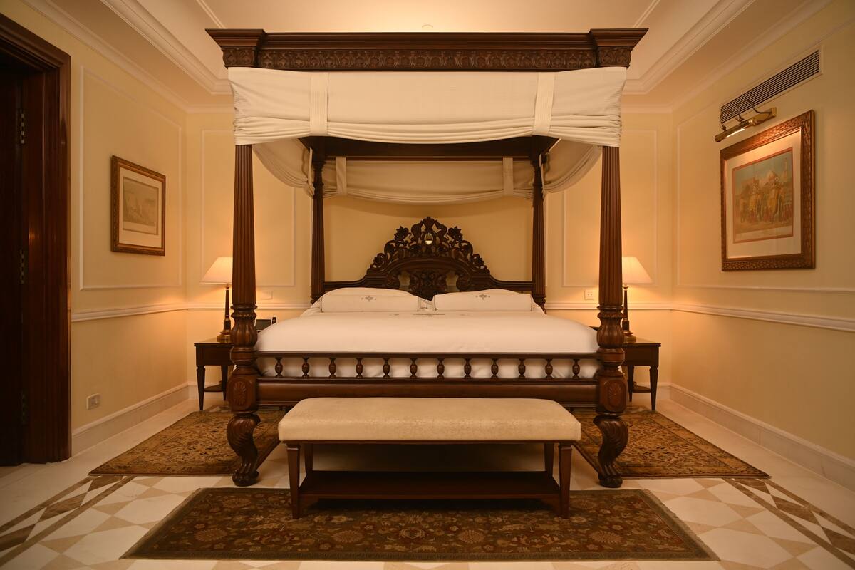 Uma cama com dossel e cortina, acompanhada de um banquinho, duas mesas de cabeceira e um tapete estampado.
