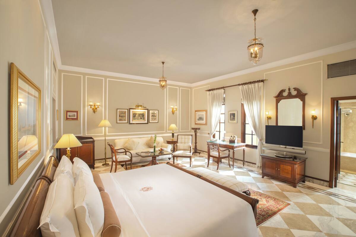 Quarto de hotel com uma cama grande, televisão, escrivaninha e área de estar com cadeiras.
