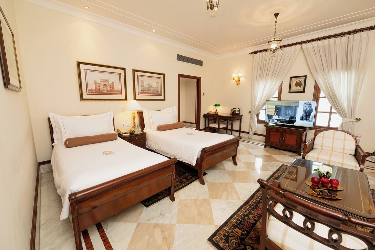 Quarto de hotel com duas camas de madeira, uma mesa de centro de vidro com frutas, uma TV e vista para um edifício pela janela.