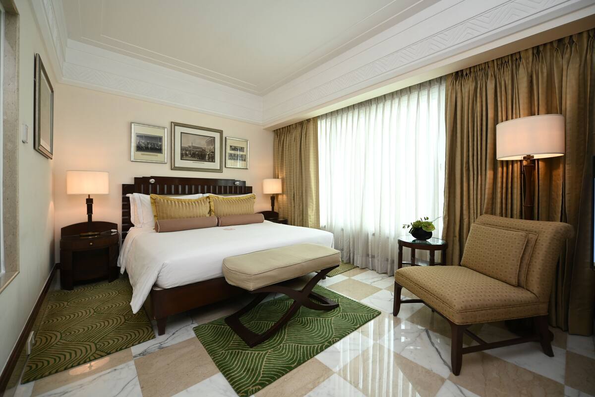 Quarto de hotel com cama, uma cadeira, mesa de cabeceira, abajur e janela com cortinas.