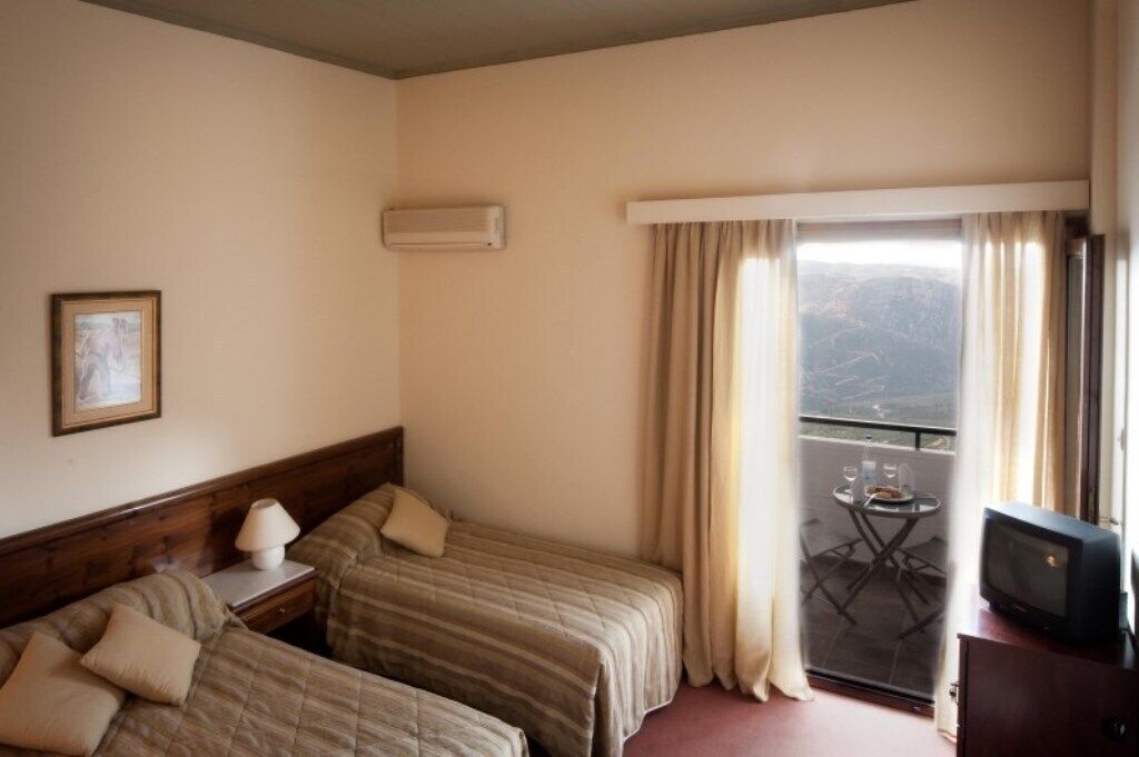 Quarto de hotel com duas camas, uma mesa pequena, televisão e varanda com vista.