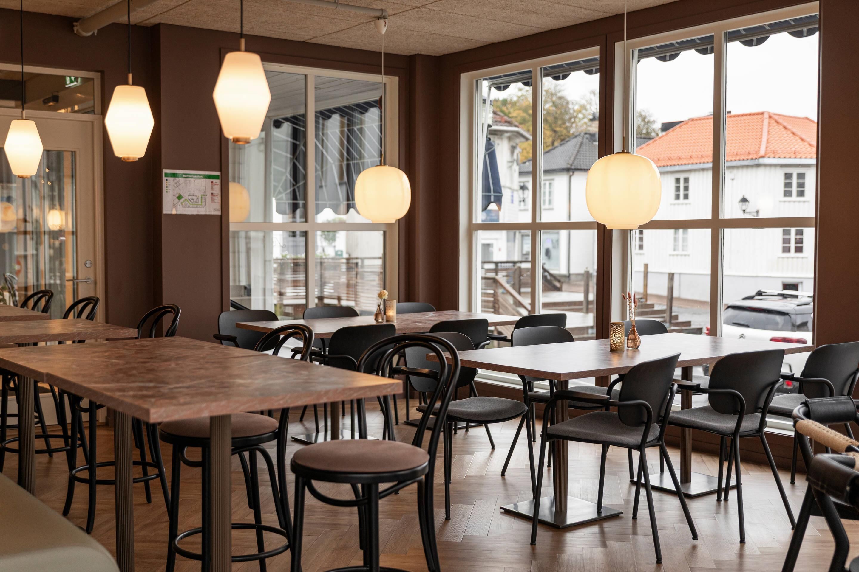 Interior de restaurante com mesas de madeira e cadeiras pretas, grandes janelas com vista para edifícios e árvores, e luminárias pendentes.