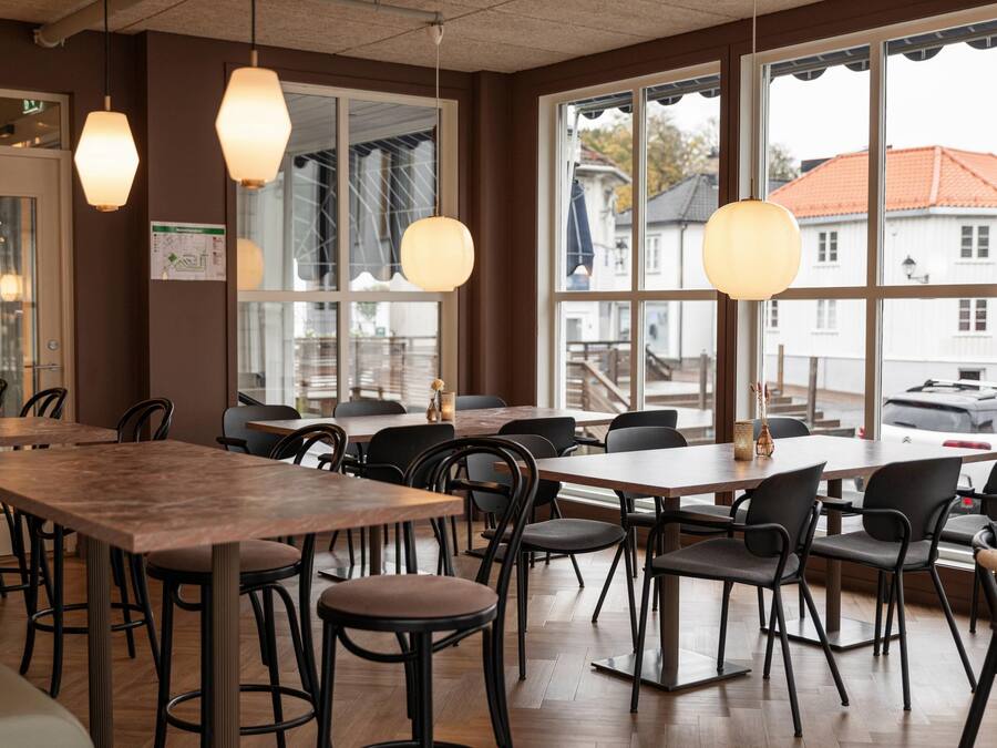 Interior de restaurante com mesas de madeira e cadeiras pretas, grandes janelas com vista para edifícios e árvores, e luminárias pendentes.