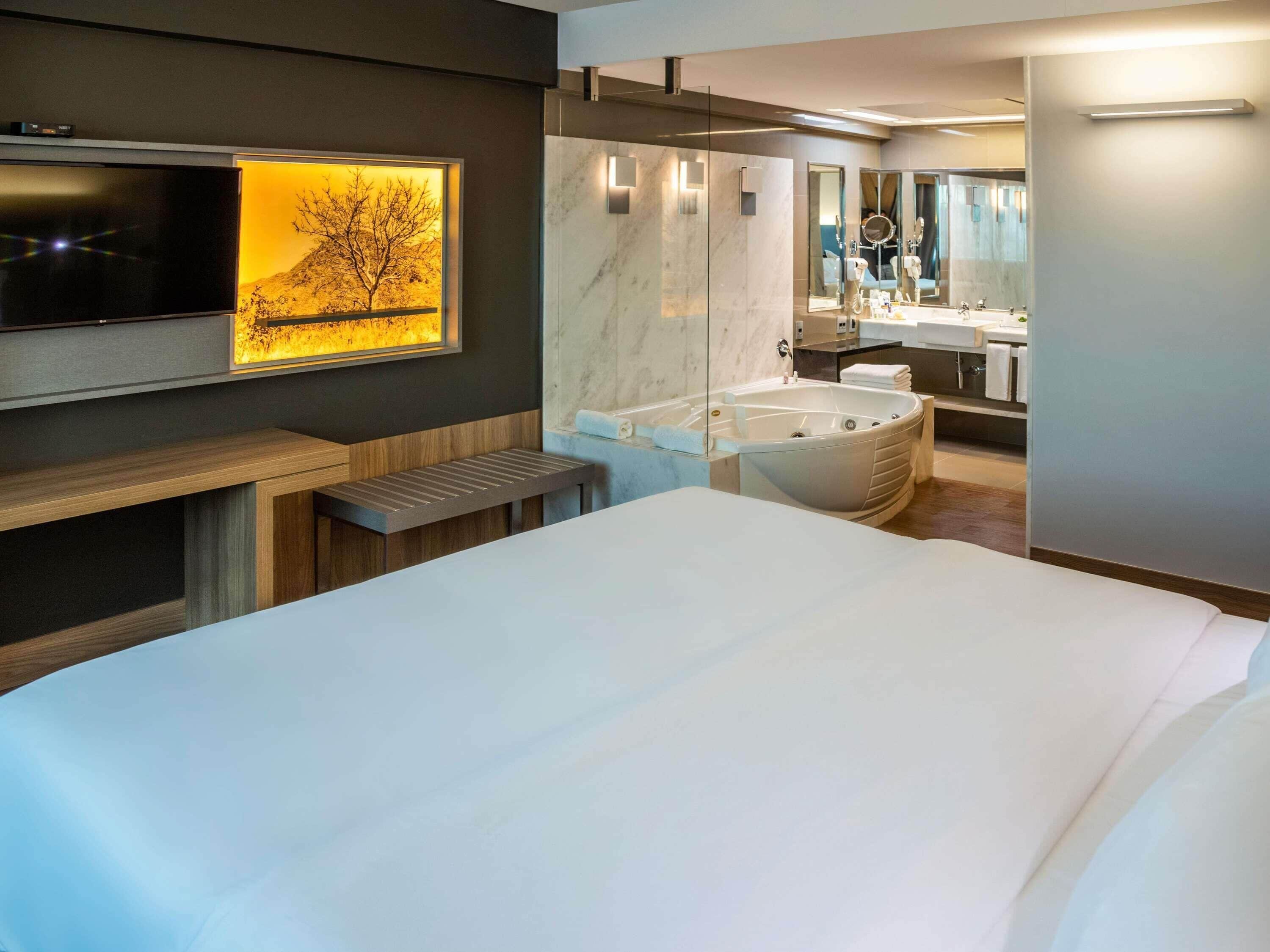 Quarto de hotel moderno com uma cama grande, televisão fixada na parede, banheira de vidro e toucador com espelho.