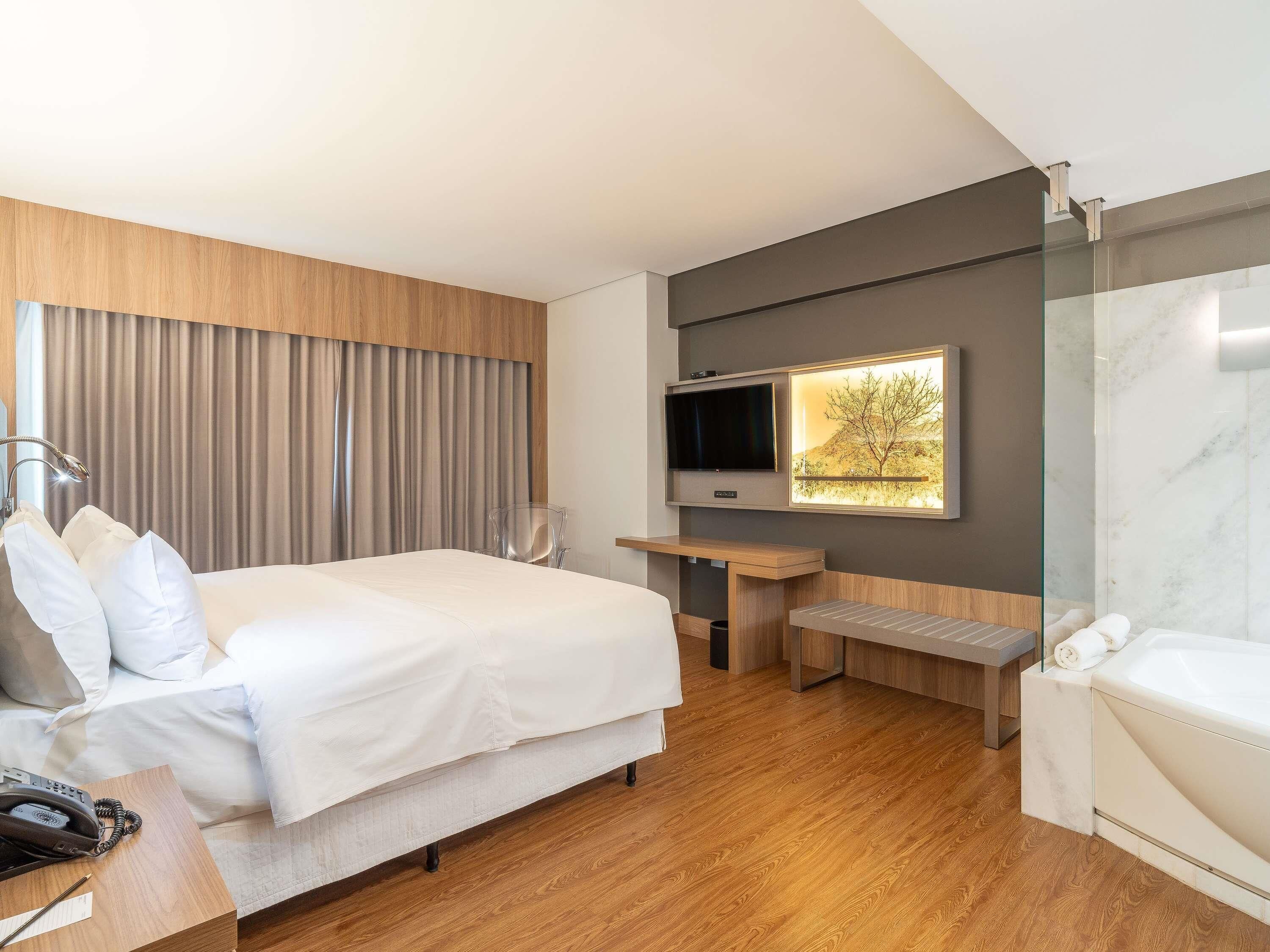 Quarto de hotel com cama, escrivaninha, televisão, chuveiro e vista para uma árvore pela janela.