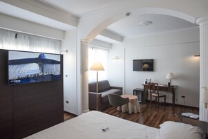Quarto de hotel com cama, escrivaninha, cadeira, sofá e televisão na parede.