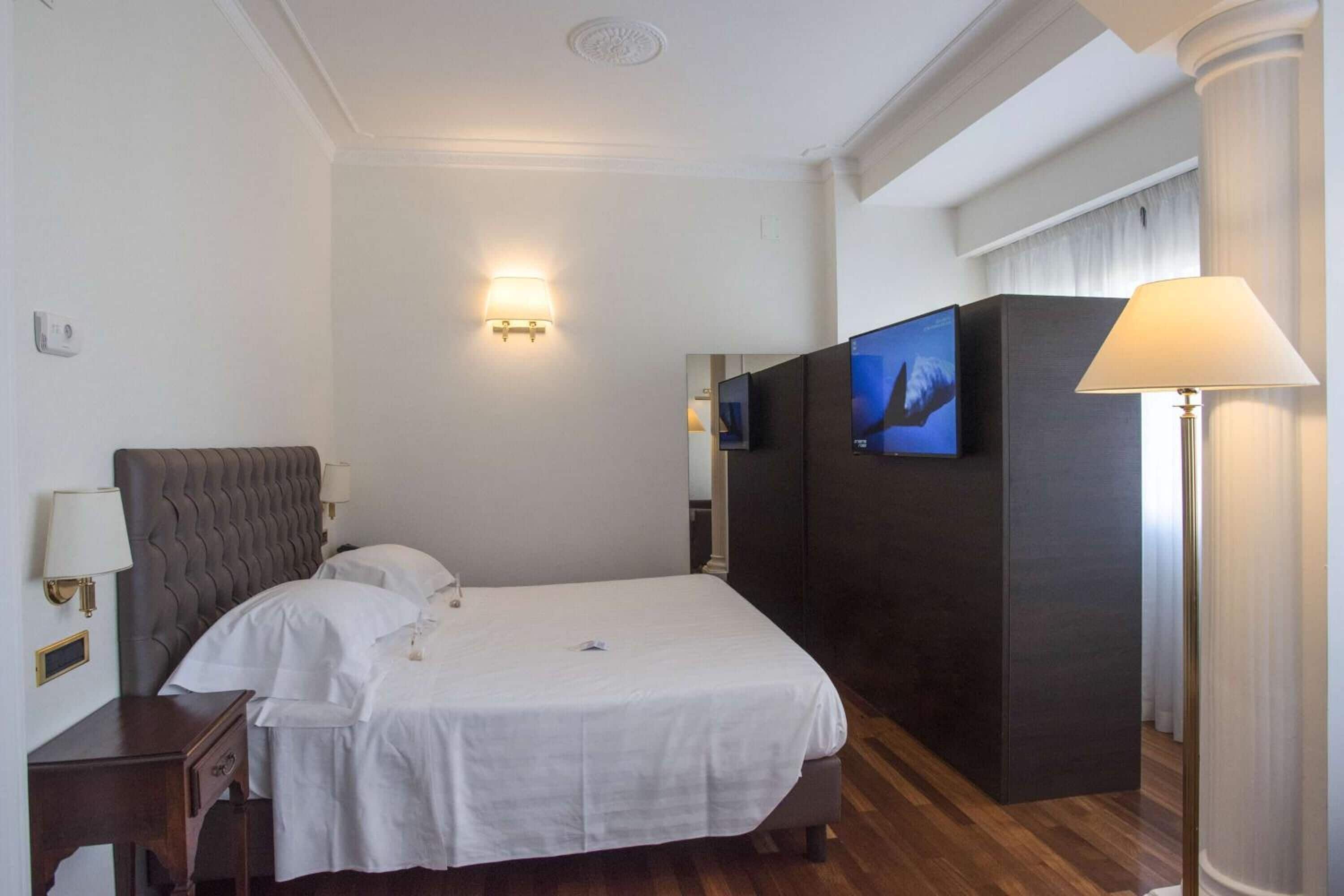 Quarto de hotel com cama, abajures, televisão e uma luminária em uma mesinha.