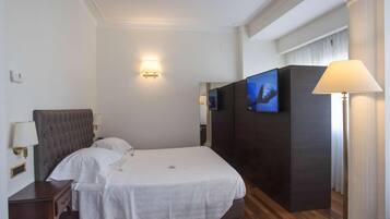 Quarto de hotel com cama, abajures, televisão e uma luminária em uma mesinha.