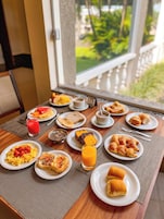 Mesa de café da manhã com diversos pratos, incluindo ovos mexidos, torradas, croissants e um copo de suco de laranja.