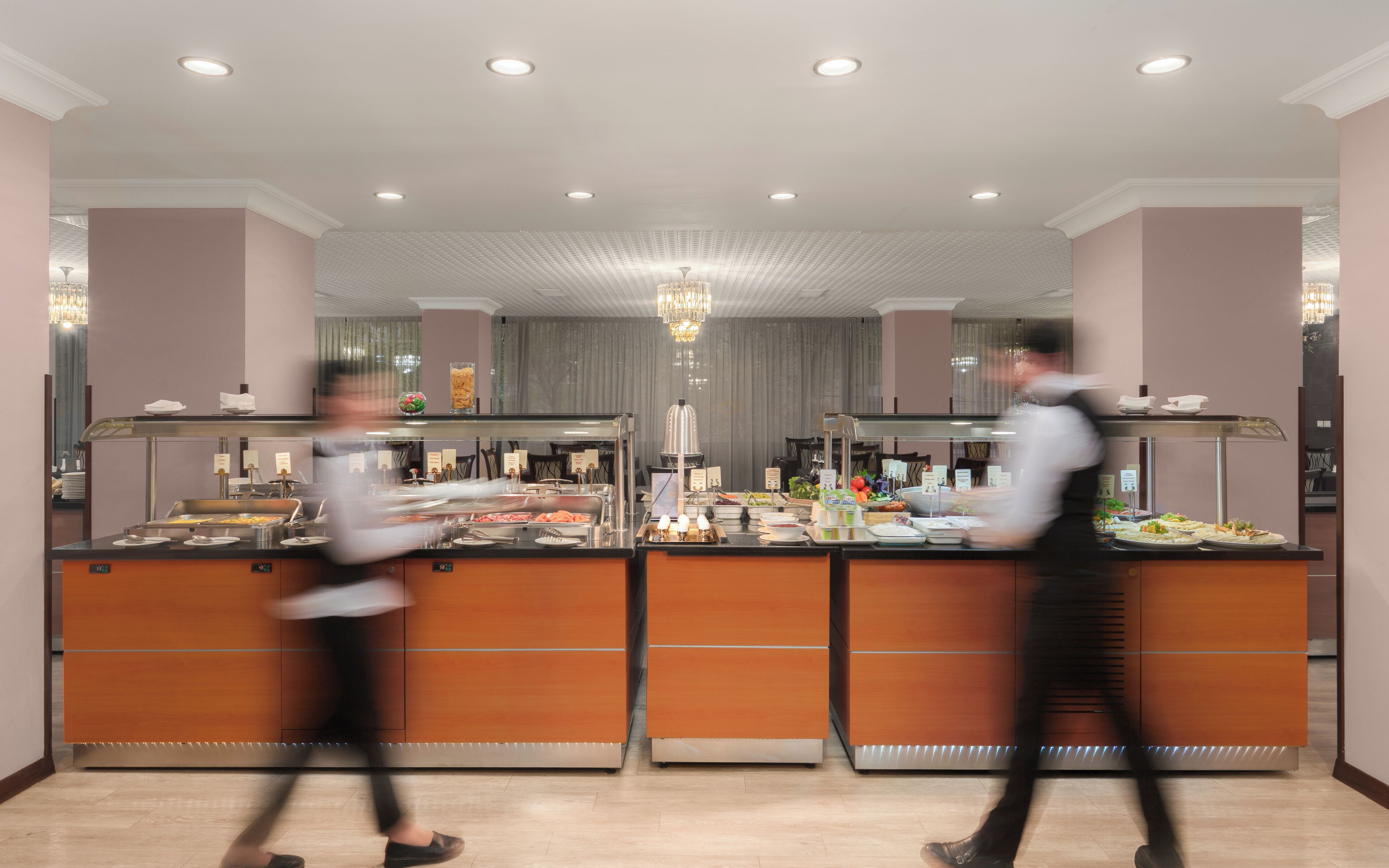 Uma área de buffet com várias estações de comida, um candelabro central e uma parede espelhada.