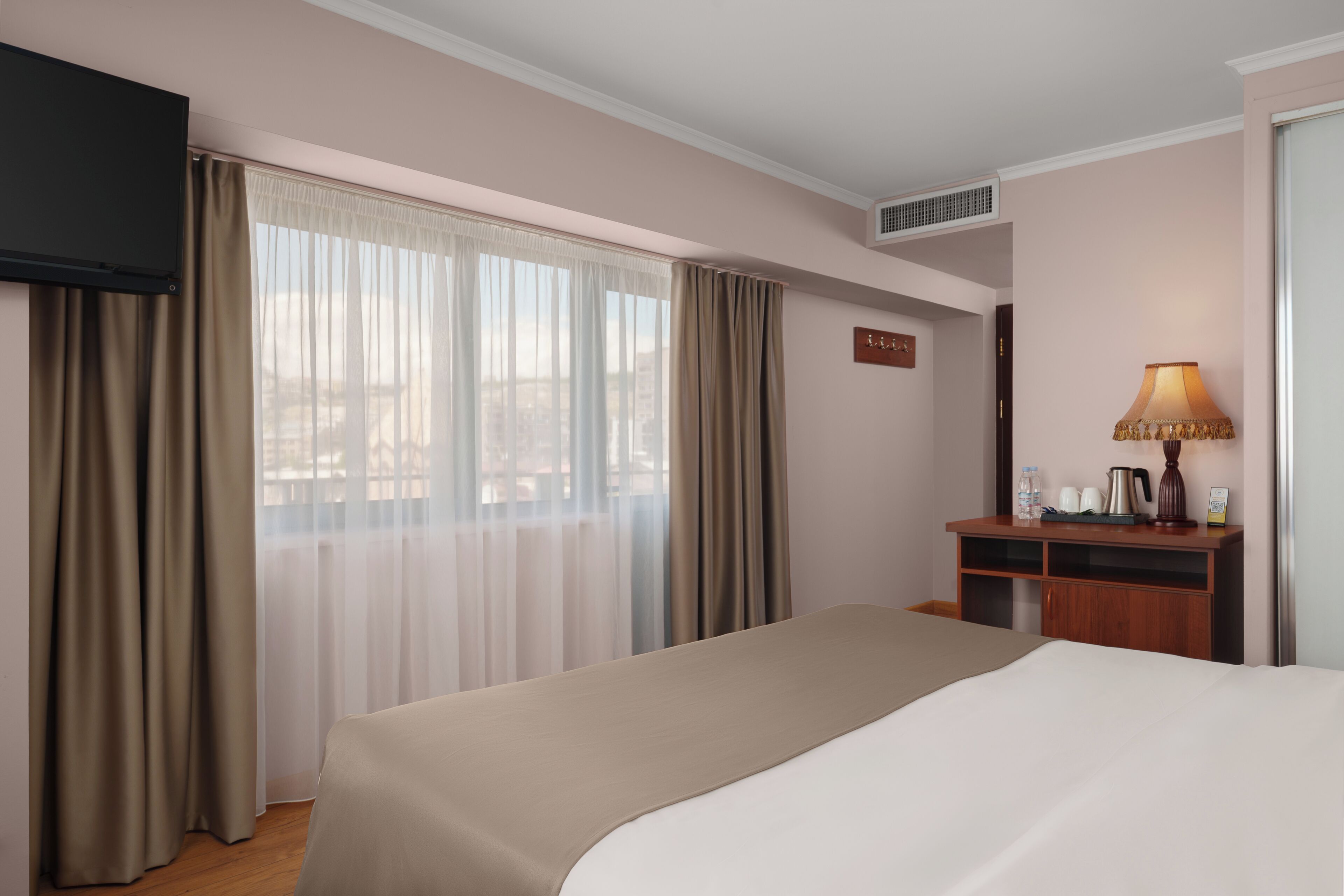 Quarto de hotel com cama, escrivaninha com luminária, televisão e vista da cidade através de grandes janelas.