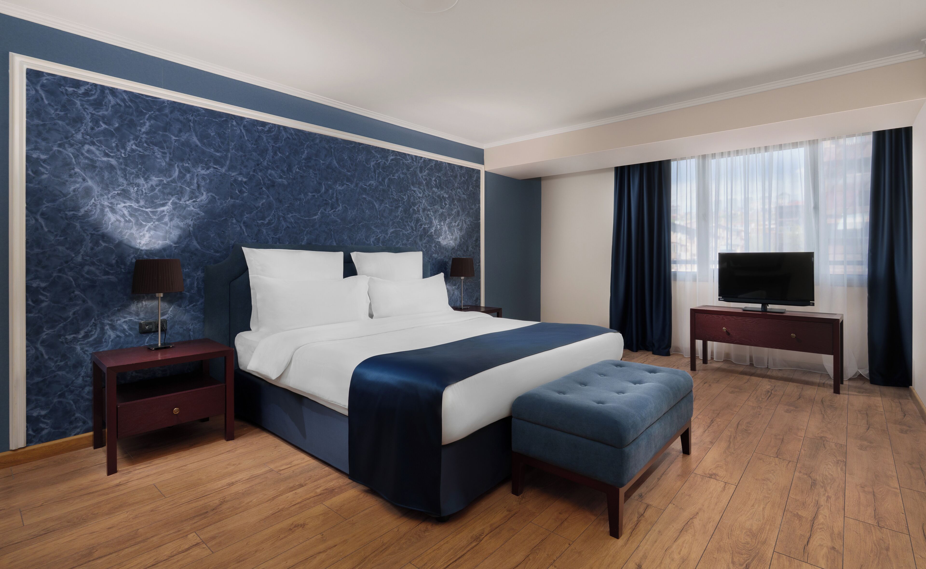 Quarto de hotel com cama, televisão, escrivaninha e janela com cortinas.