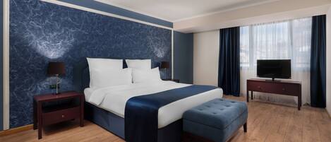 Quarto de hotel com cama, televisão, escrivaninha e janela com cortinas.