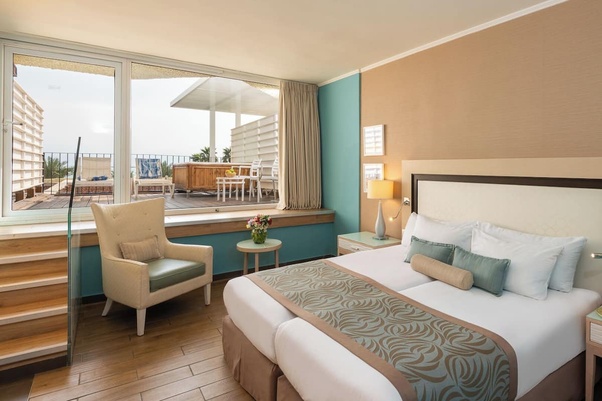 Quarto de hotel com cama grande, uma cadeira, uma mesinha e vista para o exterior.