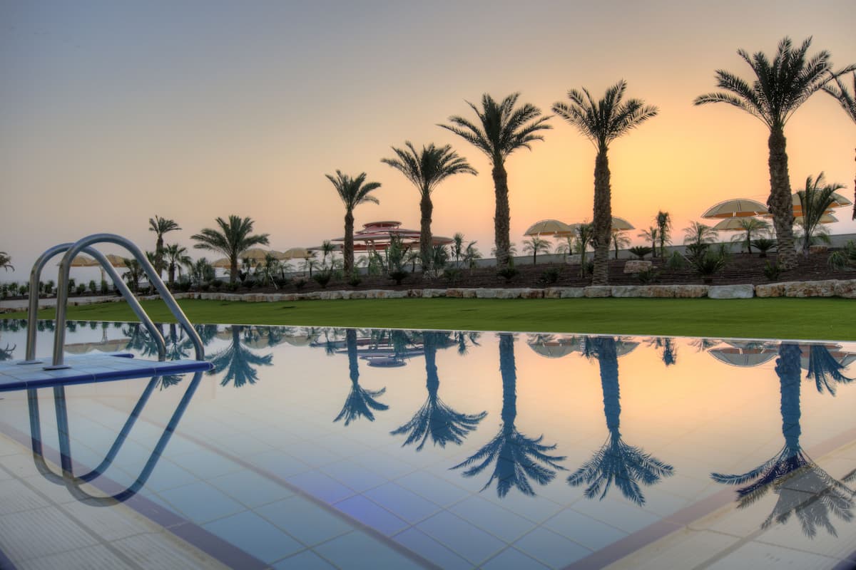 Piscina com reflexo nítido de palmeiras e um pôr do sol ao fundo.