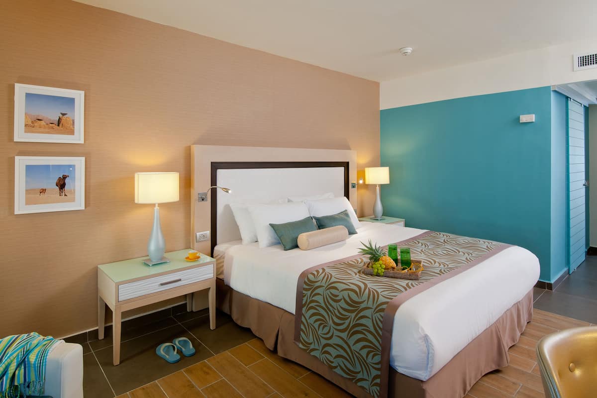 Quarto de hotel com uma cama grande, duas mesas de cabeceira, um criado-mudo, uma cadeira e uma porta que dá para outro quarto.