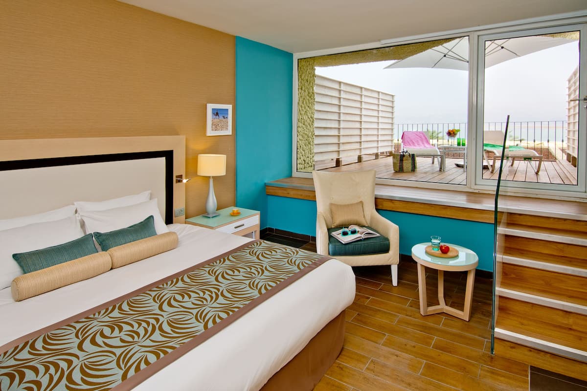 Quarto de hotel com uma cama grande, uma cadeira, uma mesinha e janela com vista para a praia.
