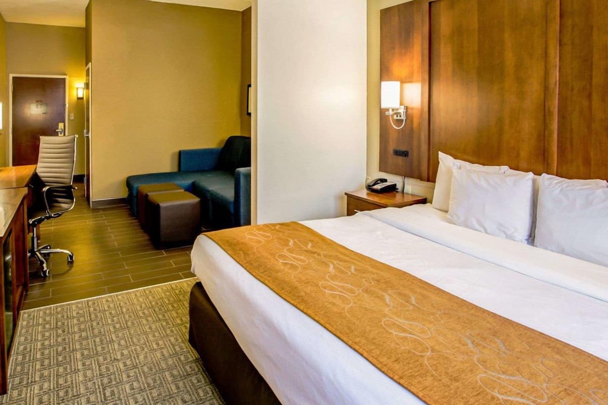 Quarto de hotel com uma cama grande, uma mesa com cadeira, um sofá e uma pequena mesa com luminária.