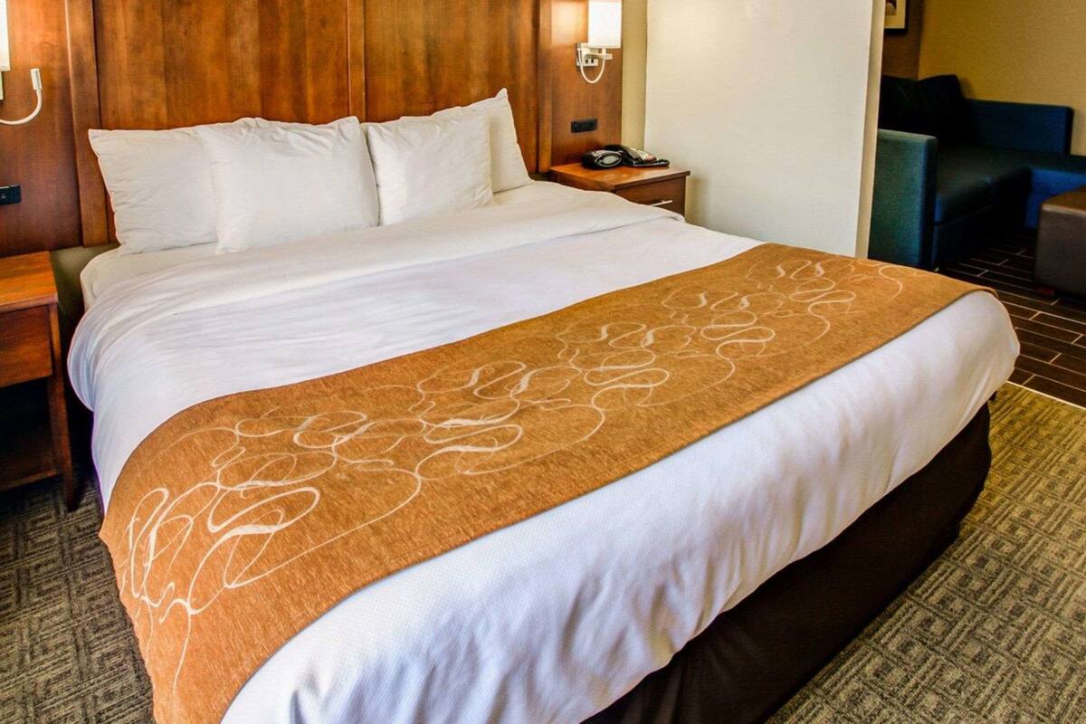 Quarto de hotel com uma cama grande, cabeceira de madeira, luminárias laterais, um telefone e uma cadeira no canto.