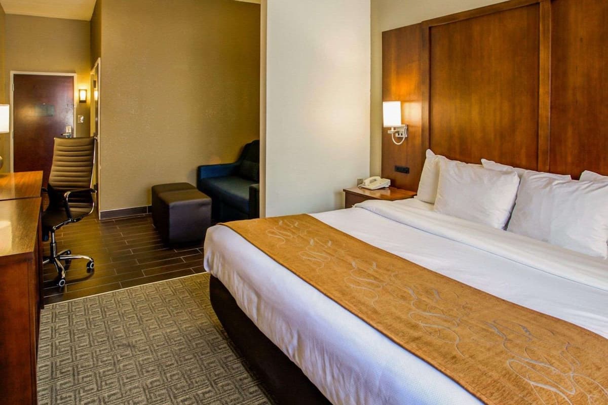 Quarto de hotel com uma cama grande, uma escrivaninha com cadeira, uma mesinha e uma mesa de cabeceira com abajur.