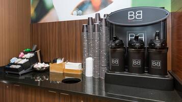 Uma área de recepção de hotel com painel de parede de madeira, balcão com pratos empilhados, guardanapos e um espaço com café, incluindo dispensadores da marca.