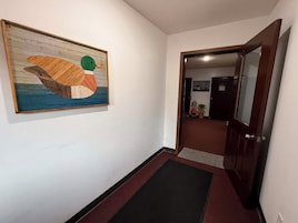 Um corredor com um quadro de um pato de madeira, um tapete escuro na entrada e uma porta que dá para um quarto com uma planta e uma pequena estátua.