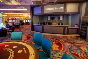 Uma área de casino com máquinas de slot, uma mesa de roleta e um balcão de recepção com a inscrição "CASINO HOST".