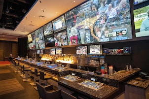 Uma área de bar com várias telas exibindo eventos esportivos, um balcão comprido com banquetas e um teto bem iluminado com luminárias embutidas.