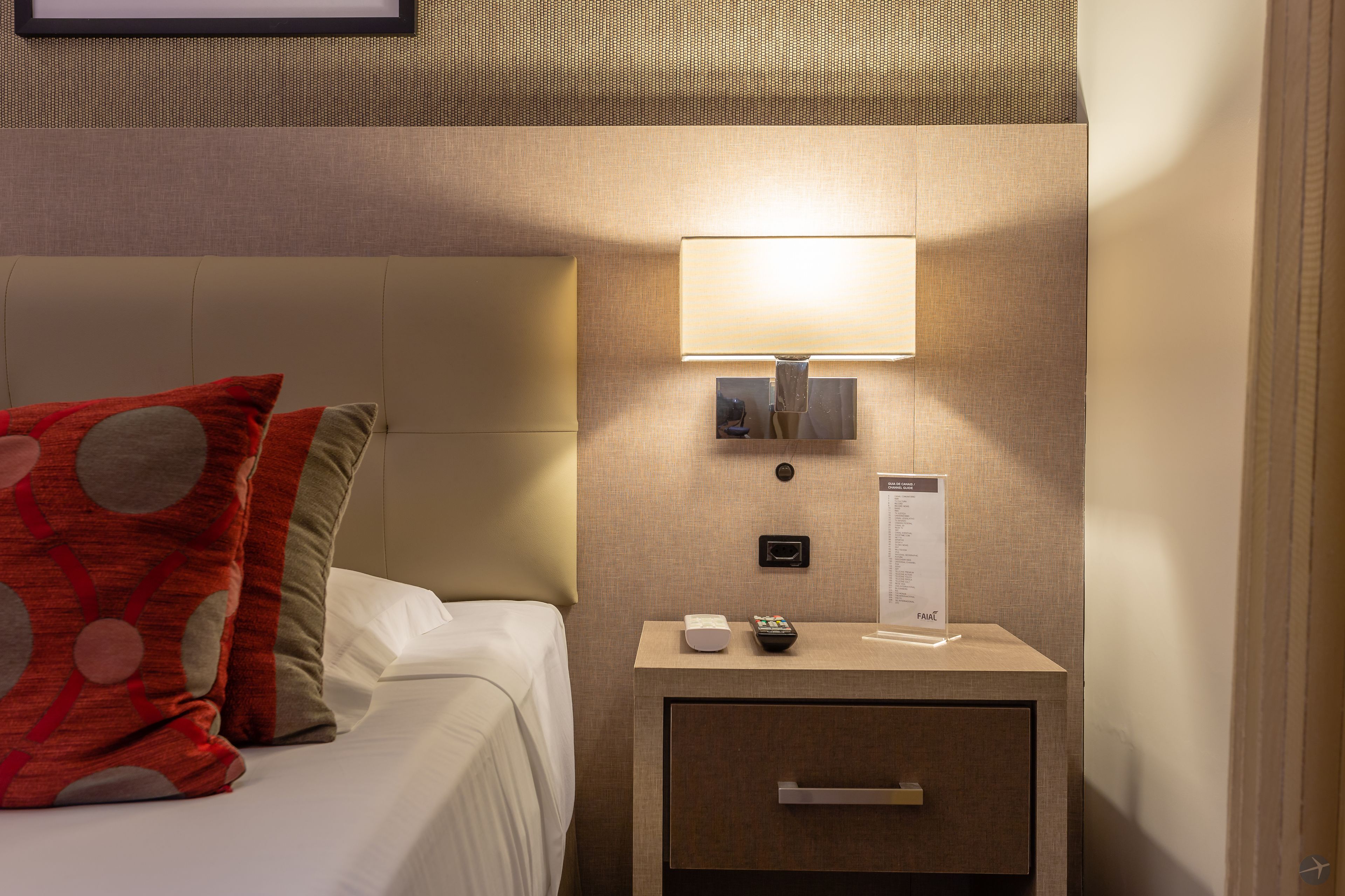 Quarto de hotel com uma cama, mesa de cabeceira com luminária, controle remoto e suporte para menu.