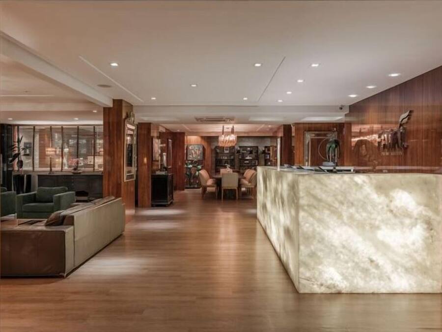 Um hall de hotel moderno com um balcão de recepção de mármore, painéis de madeira e uma área de estar com sofás e uma mesa de jantar.